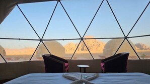 Quarto com uma cama, duas cadeiras e uma mesa pequena. O quarto possui uma janela grande com vista para montanhas.