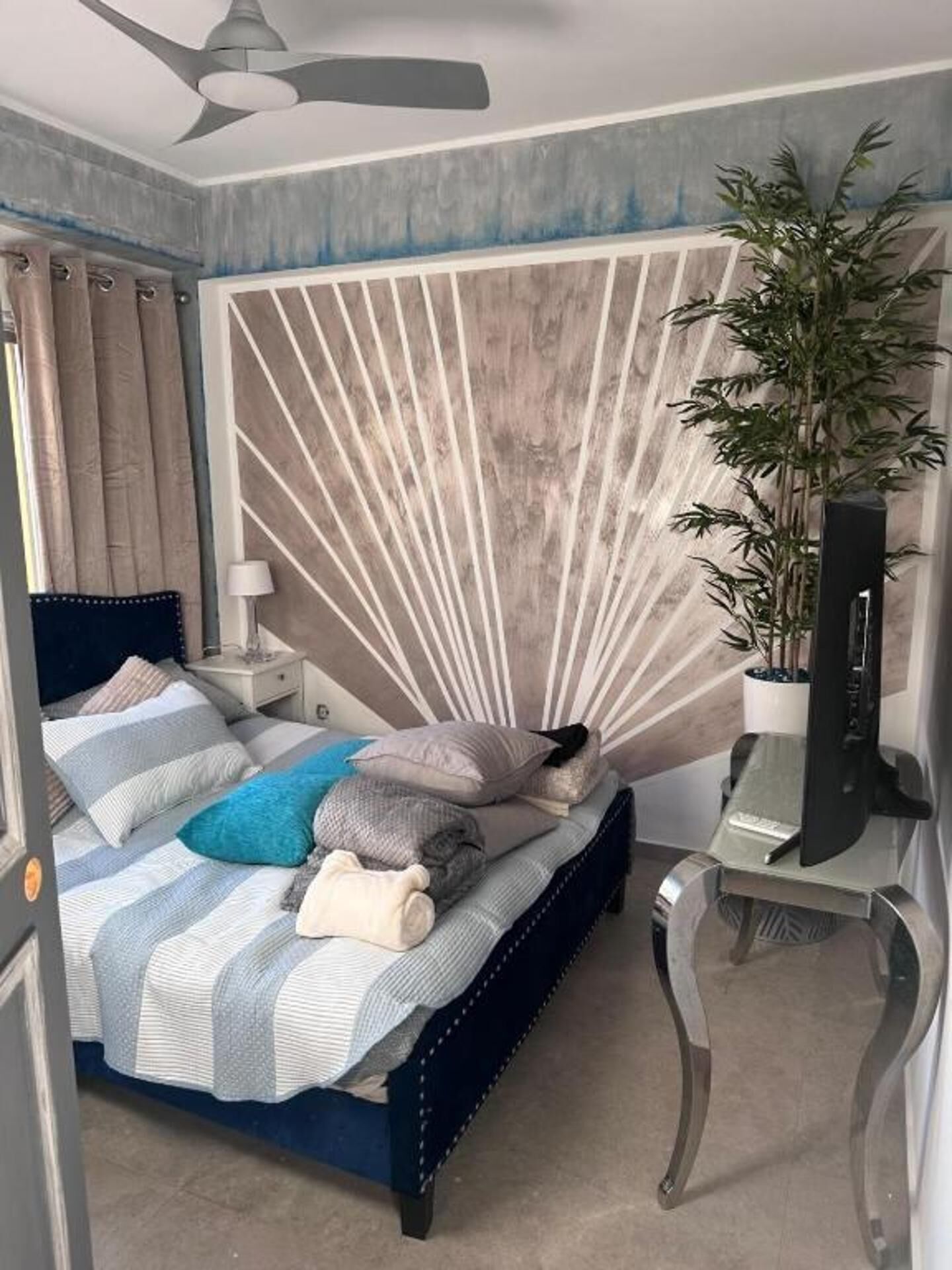 Quarto com cama, mesa de cabeceira, luminária, ventilador de teto e planta em vaso.