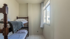 Quarto pequeno e estreito com uma cama de solteiro, cabeceira de madeira e uma janela com cortinas.