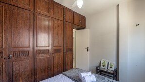 Quarto com um armário de madeira grande, uma cama com colchão cinza e uma mesinha com duas fotos emolduradas.