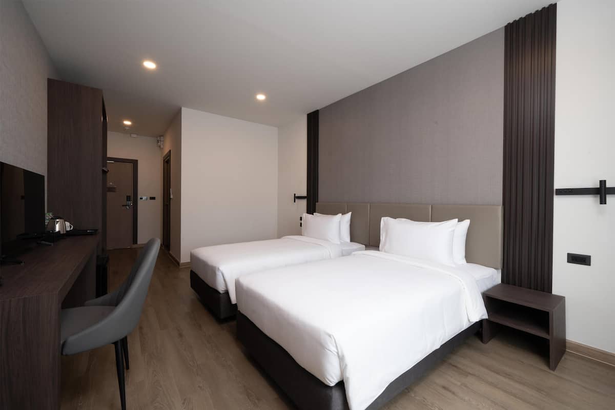 Quarto de hotel moderno com duas camas de solteiro, uma escrivaninha com cadeira e uma janela grande.