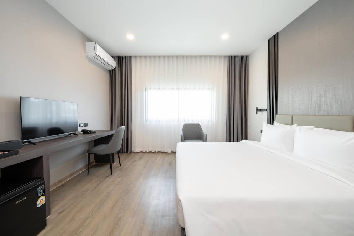 Quarto de hotel com uma cama, uma escrivaninha com televisão, uma cadeira e uma janela com cortinas.