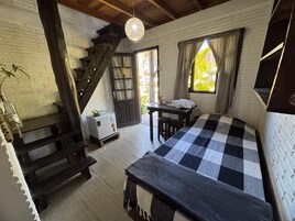 Quarto com cama xadrez, mesa pequena, geladeira, escada e janela com cortinas.