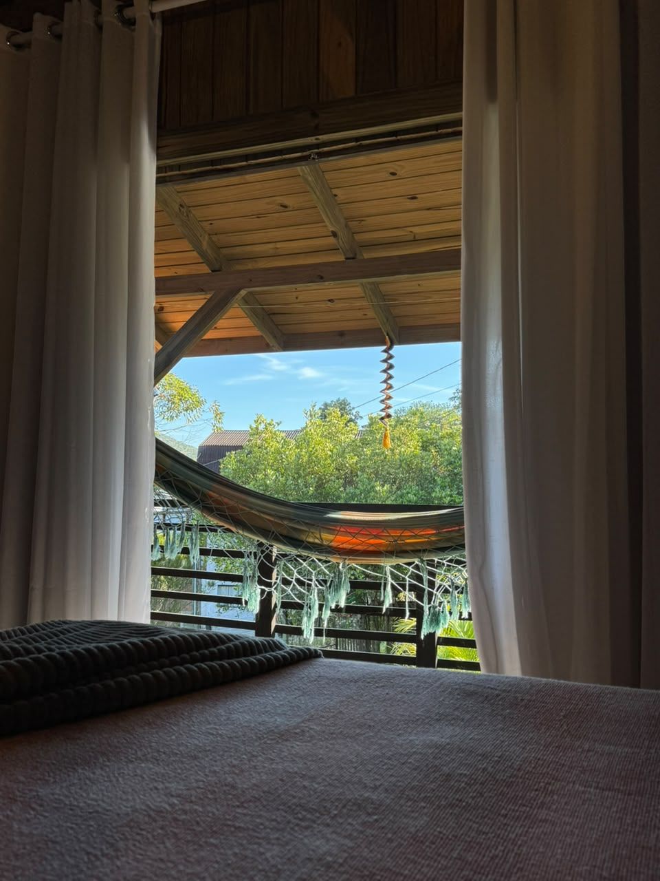 Uma rede sob teto de madeira, vista para o verde lá fora e uma cama com travesseiro.