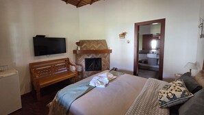 Um quarto com uma cama, um banco de madeira, uma TV fixada na parede, uma lareira e um banheiro visível por uma porta aberta.