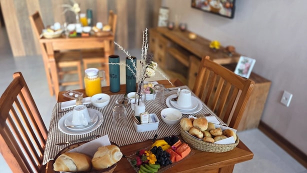 Uma mesa posta para dois com pratos, xícaras, cesta de pastelaria e uma saladeira com frutas.