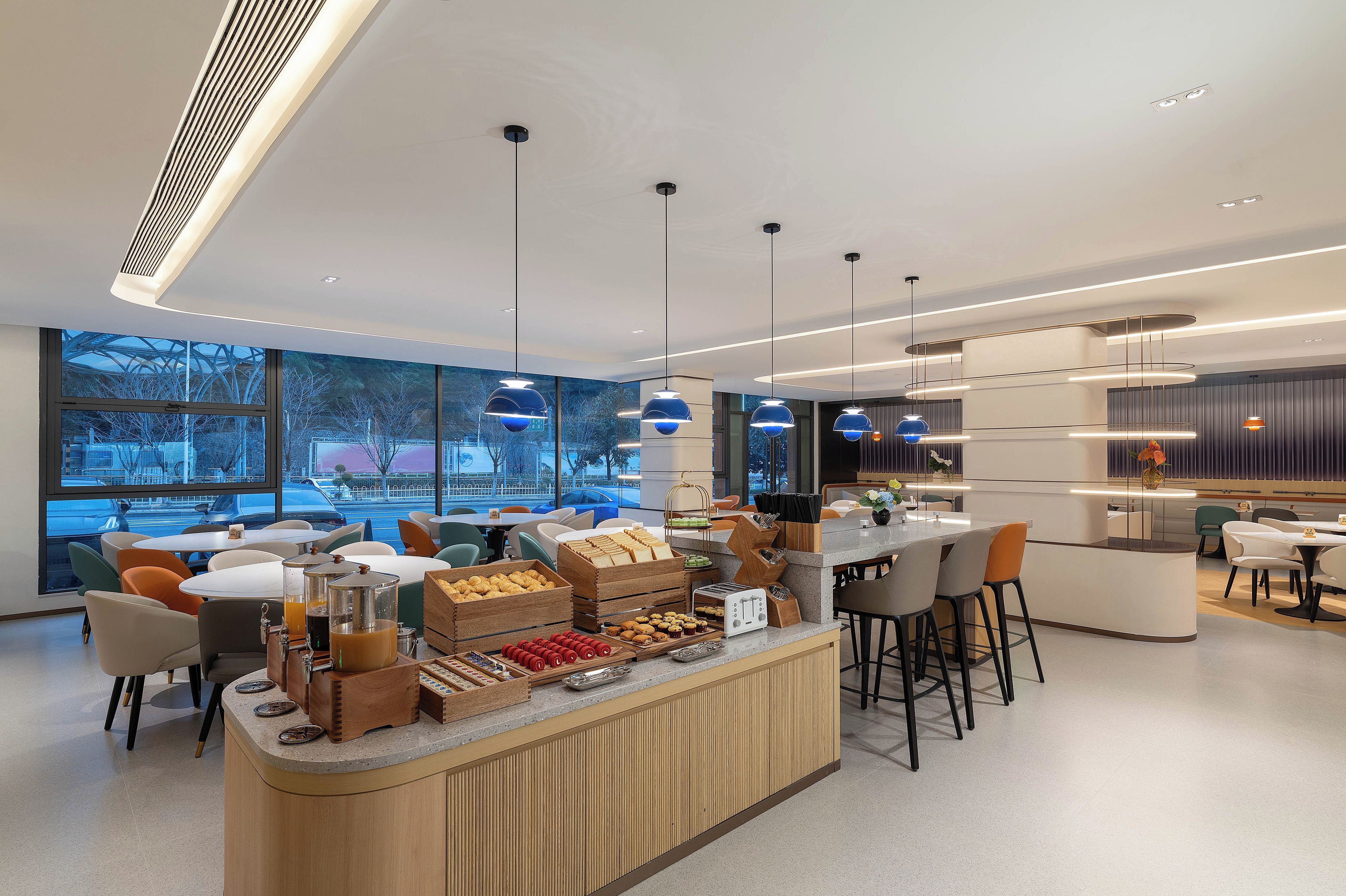 Um café moderno com um balcão central que oferece diversas opções de comida, rodeado por áreas de estar com grandes janelas que dão para o exterior.