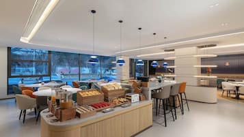 Um café moderno com um balcão central que oferece diversas opções de comida, rodeado por áreas de estar com grandes janelas que dão para o exterior.