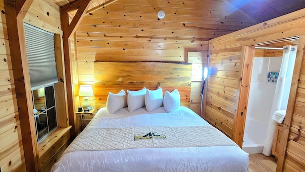Quarto de cabana de madeira com uma cama grande, um banquinho, uma janela com persianas e teto de madeira.