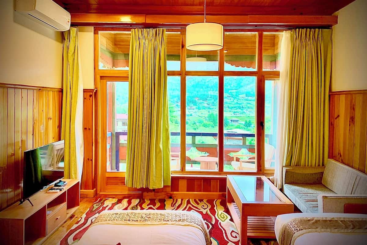 Quarto com cama, televisão, mesa e sofá. Cortinas e janela com vista para áreas verdes.