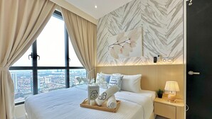 Quarto de hotel com uma cama grande, uma mesa de cabeceira com abajur, vista da cidade pela janela e um painel floral na parede.