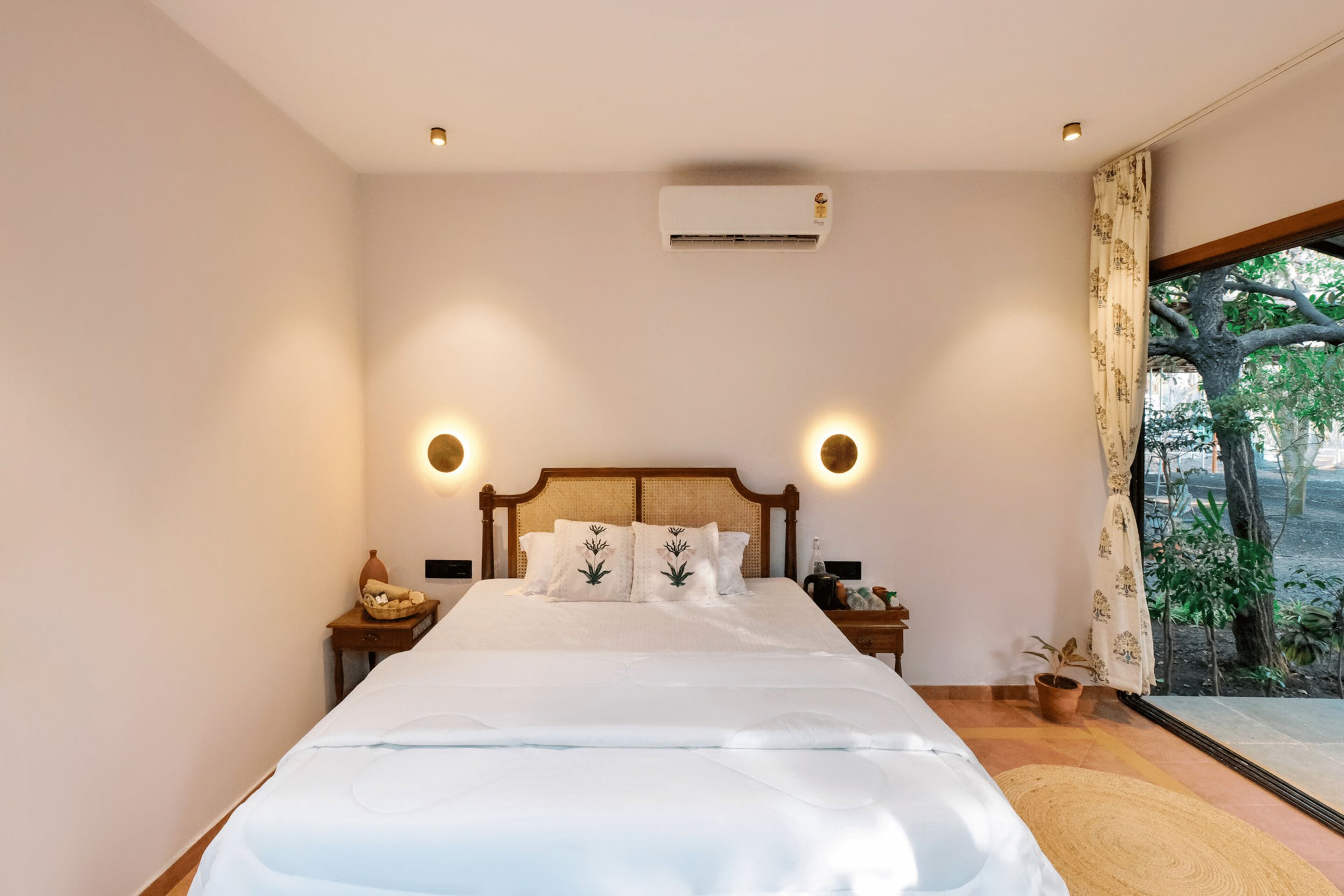 Quarto com cama, mesas de cabeceira, ar condicionado e vista para o verde pela janela.