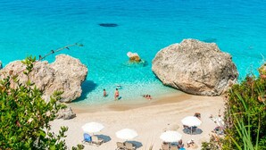 Uma praia com águas turquesa cristalinas, pedras grandes e pessoas nadando. Há cadeiras de praia e guarda-sóis.