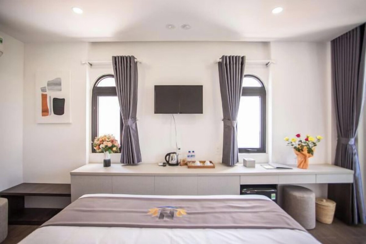 Quarto de hotel moderno com cama, escrivaninha com TV de tela plana, duas janelas com cortinas, uma mesinha com um vaso de flores e uma TV fixada na parede.