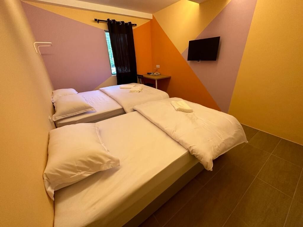 Quarto de hotel com duas camas, uma escrivaninha pequena, televisão fixada na parede e uma janela com cortinas.