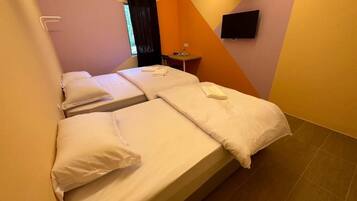 Quarto de hotel com duas camas, uma escrivaninha pequena, televisão fixada na parede e uma janela com cortinas.