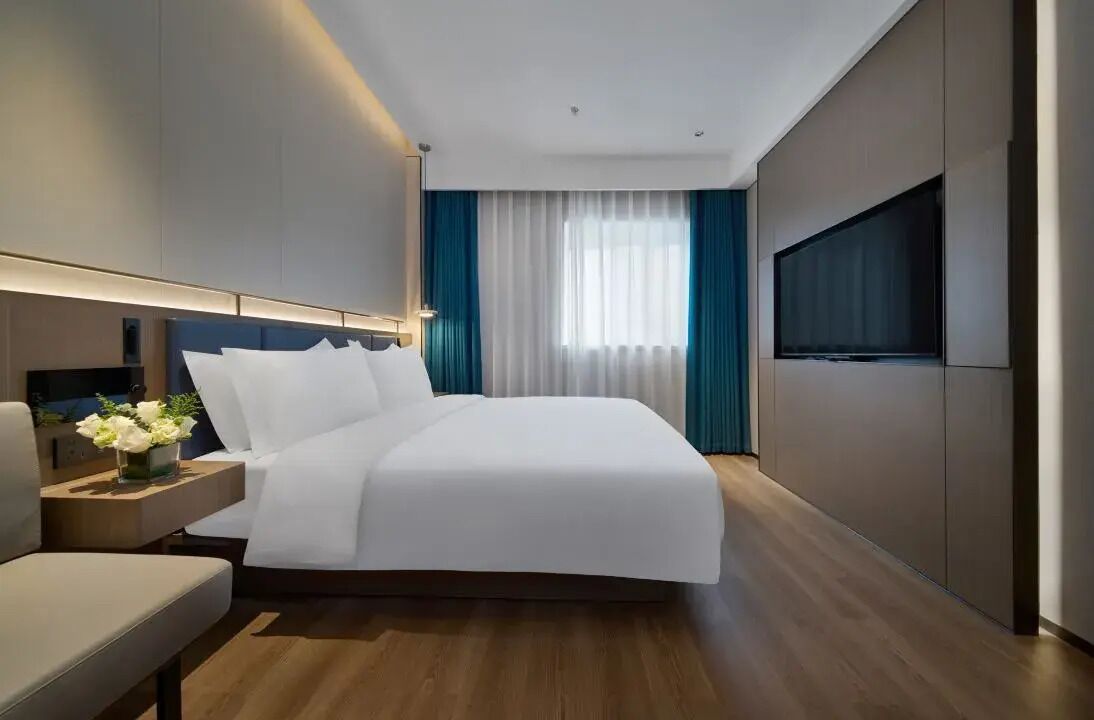 Quarto de hotel moderno com uma cama grande, televisão de tela plana, mesa de cabeceira com um vaso de flores e vista da cidade por grandes janelas.