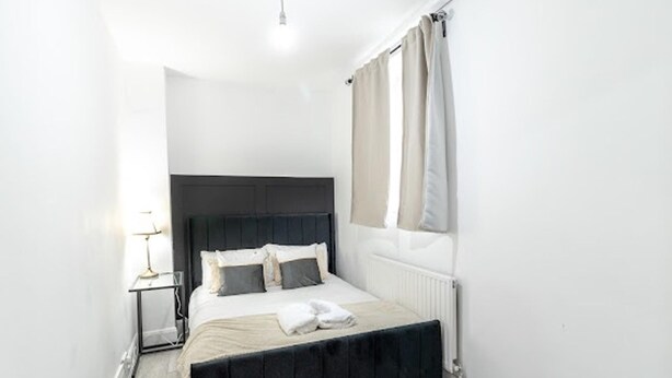 Quarto com cama, mesa de cabeceira, abajur e janela com cortinas.