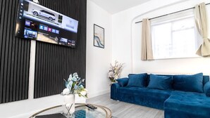Uma sala de estar moderna com um sofá azul, uma mesa de centro de vidro, uma TV montada e uma janela com cortinas.
