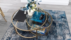 Uma mesa de vidro redonda com estrutura dourada, um caderno preto, um vaso com flores e um dispositivo eletrônico azul sobre um tapete com padrão azul.