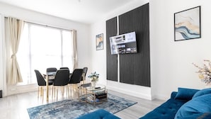 Uma sala de estar moderna com um sofá azul, uma área de jantar com cadeiras pretas, uma televisão de tela plana montada em um painel preto e obras de arte nas paredes.