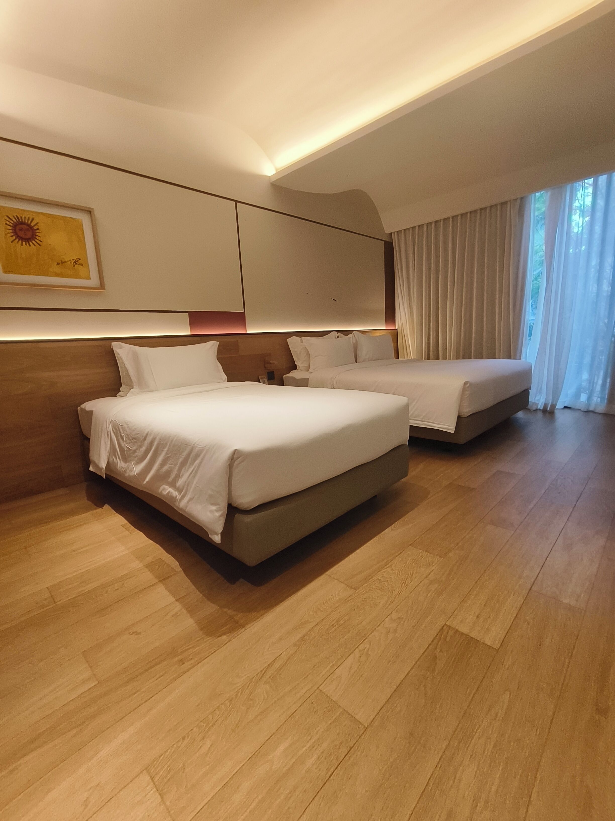 Quarto de hotel com duas camas, piso de madeira, uma janela grande com cortinas e uma obra de arte na parede.