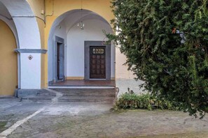 Um edifício com entrada central, uma escadaria que leva até ele e uma grande árvore à direita.