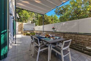 Um pátio externo com uma mesa posta para o café da manhã, cercado por cadeiras, e uma área coberta com vista para árvores e um muro de pedra.
