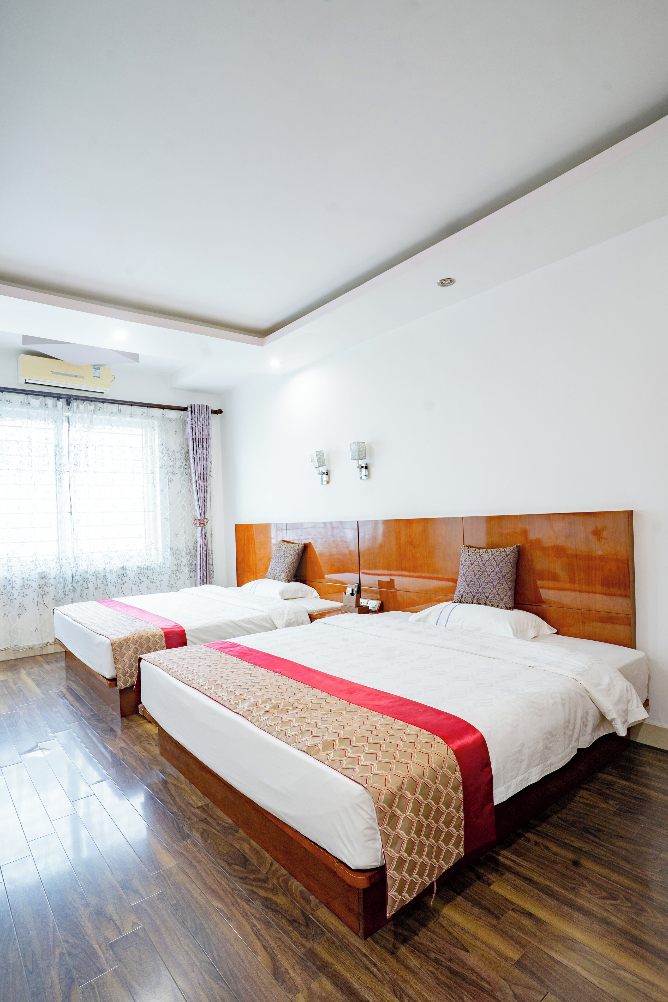 Quarto de hotel com duas camas, piso de madeira, uma janela grande com cortinas e luminária fixada na parede.