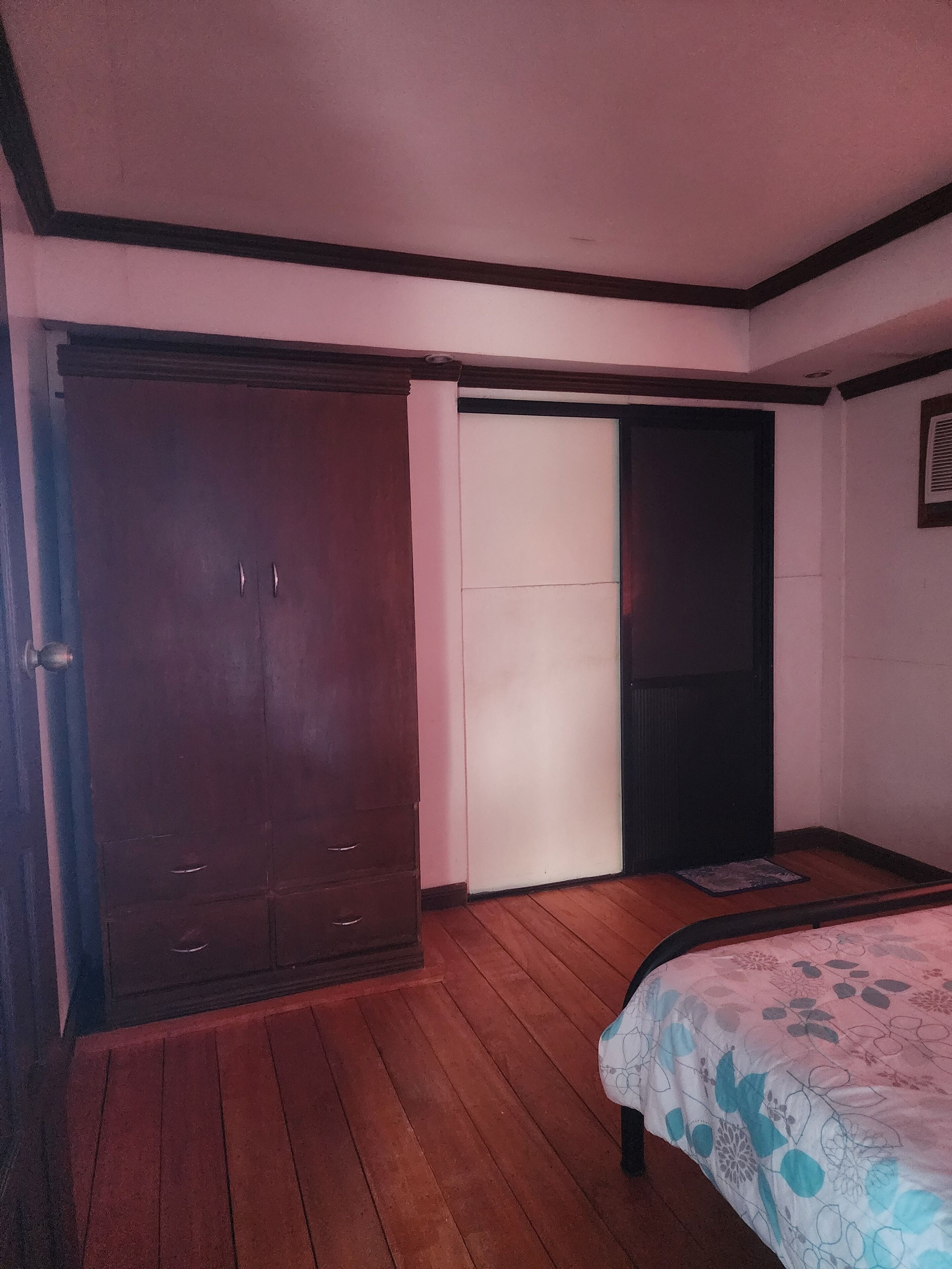 Quarto com piso de madeira, cama com colcha floral, guarda-roupa e porta para outro cômodo.