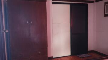 Quarto com piso de madeira, cama com colcha floral, guarda-roupa e porta para outro cômodo.