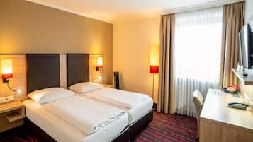 Quarto de hotel com cama de casal, luminárias laterais, mesa de trabalho com televisão e uma janela grande com cortinas.