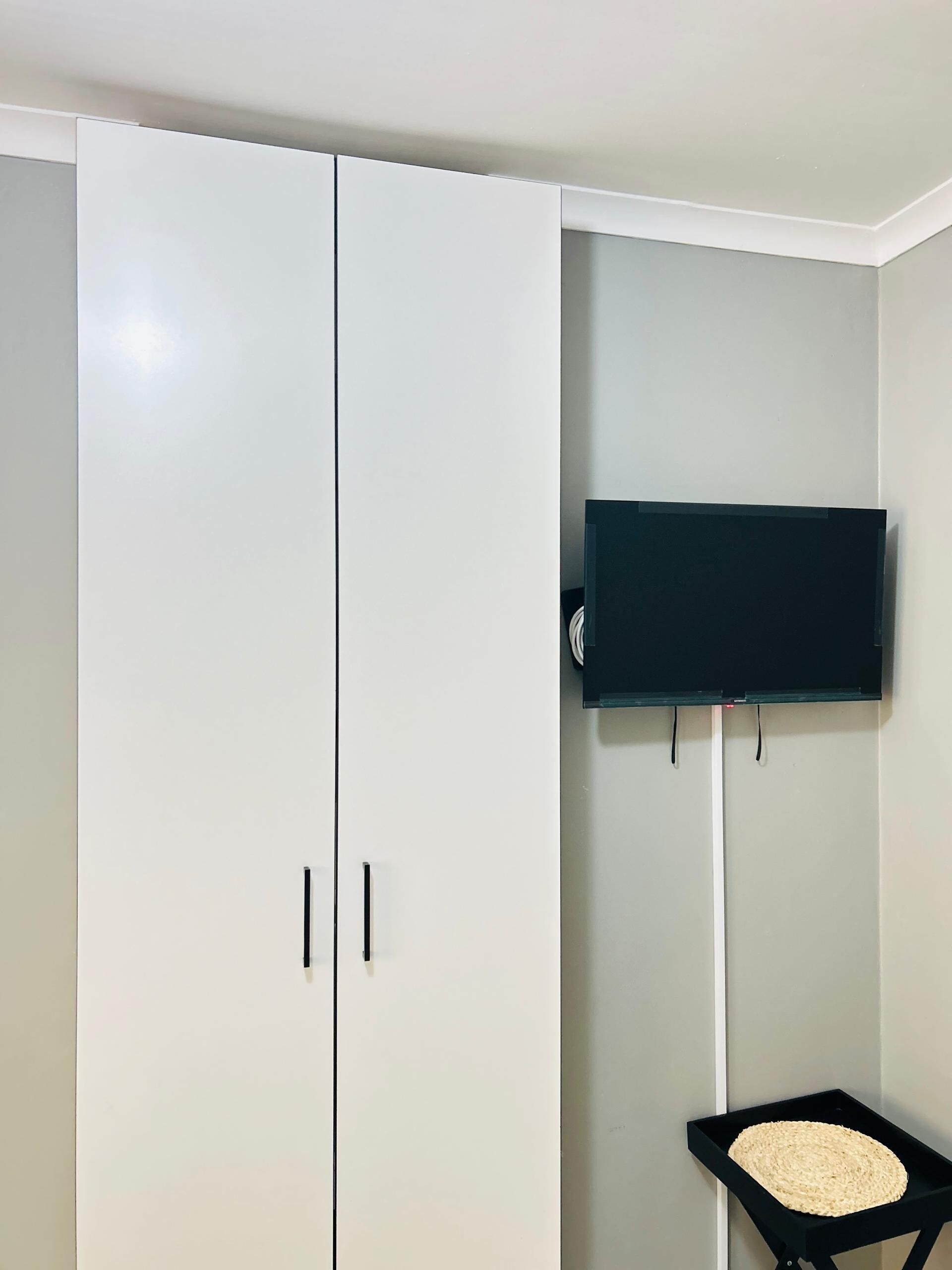 Quarto com guarda-roupa branco, televisão na parede e uma pequena mesa preta com uma cesta de vime.