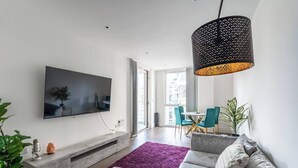 Uma sala de estar moderna com televisão de tela plana na parede, um sofá cinza com almofadas, um tapete roxo e uma luminária pendente.