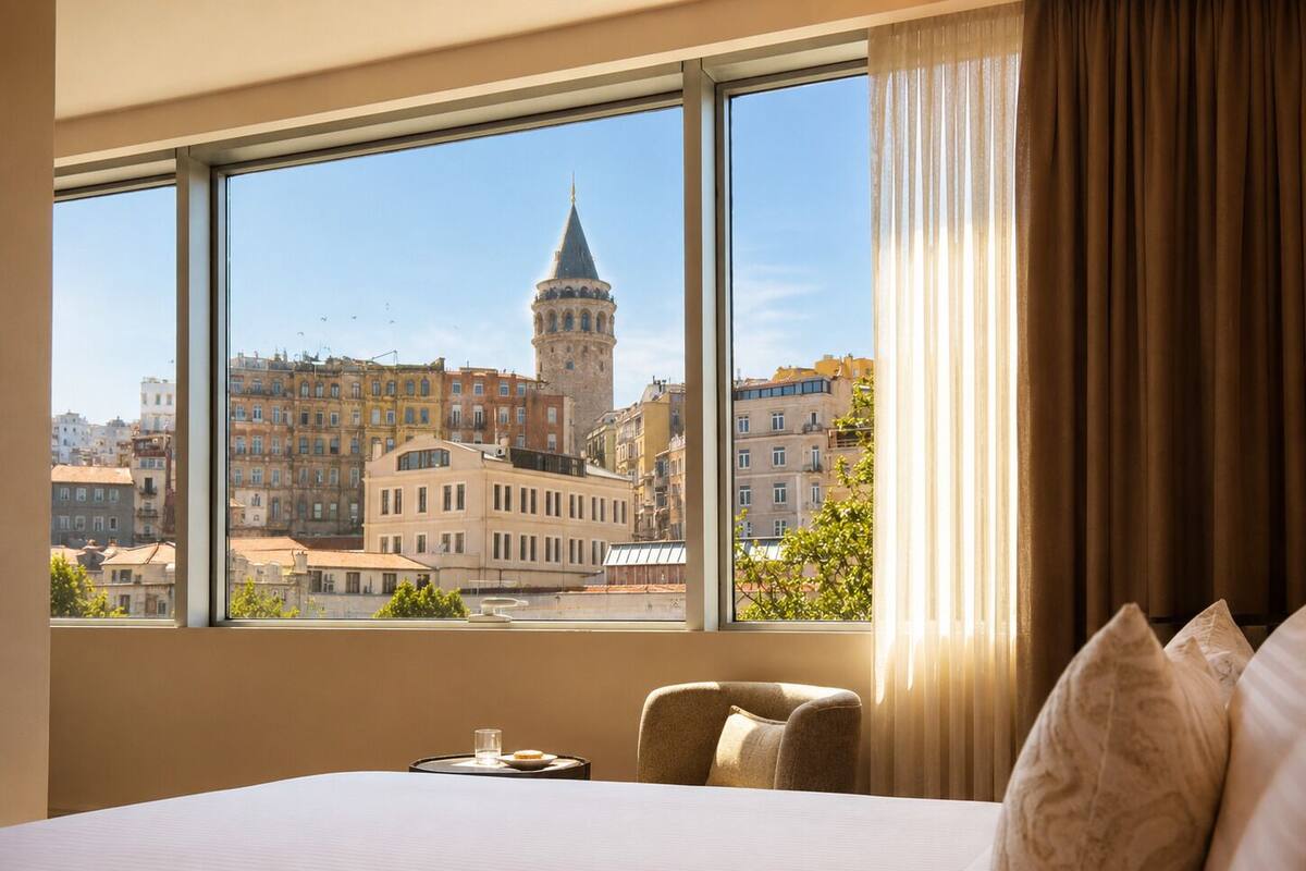 Quarto de hotel com uma janela grande, uma cama, uma cadeira e uma mesinha com um copo e uma vela.