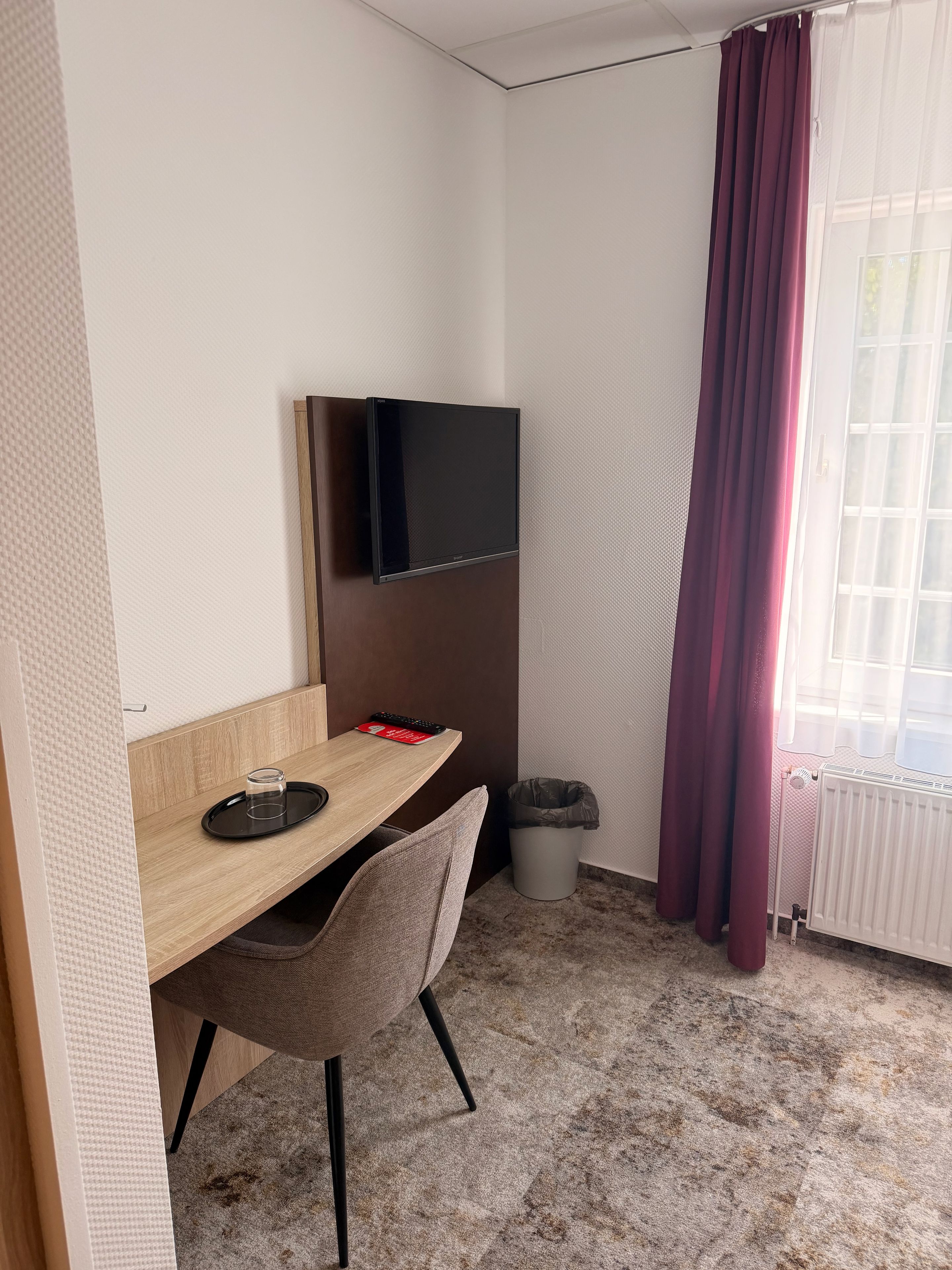 Quarto de hotel com escrivaninha de madeira, cadeira, televisão na parede, janela com cortinas e radiador.