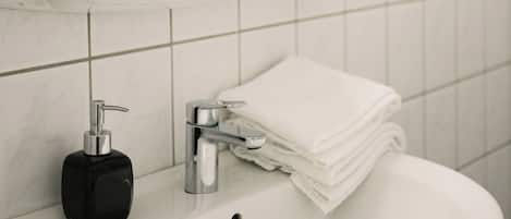 Casa de banho com lavatório branco, espelho, um copo de água, dispensador de sabonete e uma pilha de toalhas.