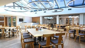 Uma ampla área de refeições interna com várias mesas e cadeiras de madeira, teto alto com claraboias circulares e uma cozinha moderna ao fundo.