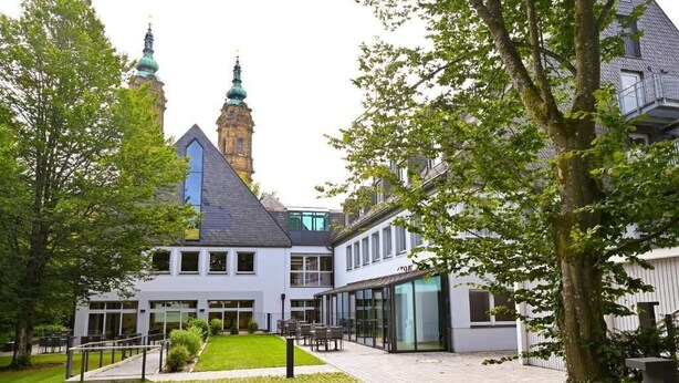Um edifício com fachada moderna, um pátio com área pavimentada e uma torre tradicional com cúpula verde ao fundo.