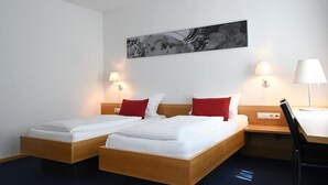 Quarto de hotel com duas camas de solteiro, uma escrivaninha, uma cadeira e uma obra de arte na parede.