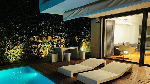 Área moderna da piscina ao ar livre, com duas espreguiçadeiras, deck de madeira e vista para o interior através de uma porta de vidro.