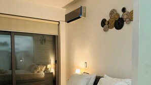 Quarto com cama, ventilador de teto, aparelho de ar condicionado, abajur e um objeto decorativo na parede.
