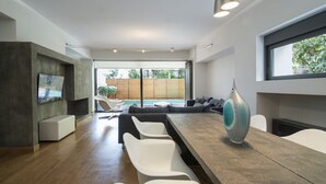 Uma sala de estar moderna com uma mesa de jantar comprida, cadeiras brancas, um sofá e uma TV montada na parede.