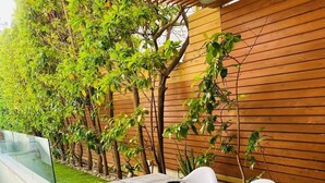 Uma área de refeições ao ar livre com mesa de concreto e cadeiras brancas, cercada por vegetação e um muro de madeira.