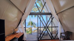 Um quarto acolhedor em formato triangular, com piso e escrivaninha de madeira, e uma cadeira. Possui uma janela grande com cortinas, oferecendo vista para palmeiras e uma praia.