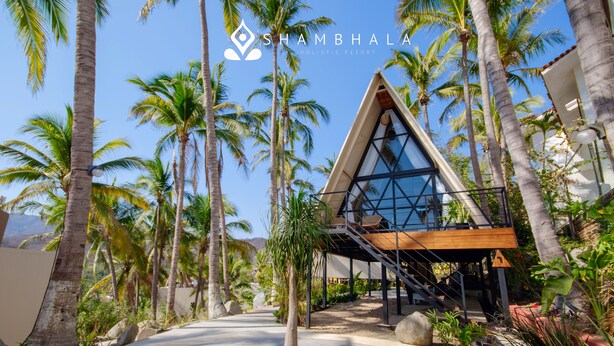 Resort com design arquitetônico moderno, apresentando uma fachada de vidro imponente e um deck de madeira. Palmeiras margeiam o caminho que leva à entrada.