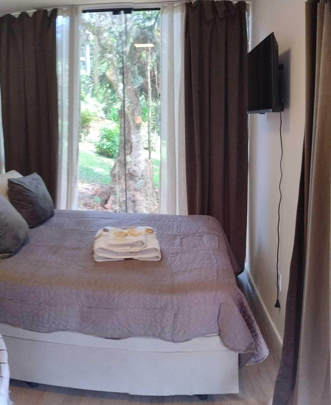Uma cama bem arrumada com uma toalha dobrada, uma televisão de tela plana na parede e uma janela com cortinas.