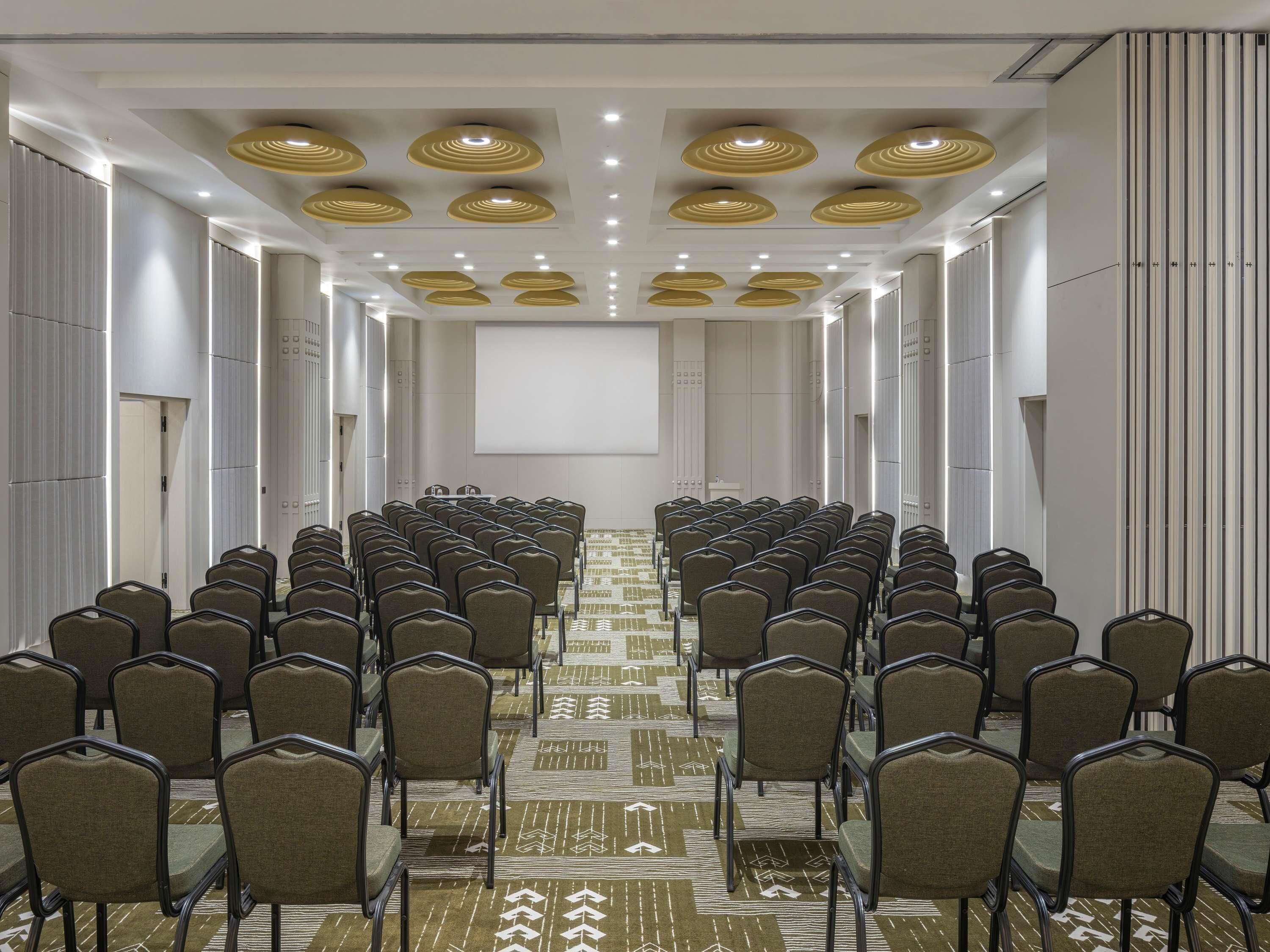 Sala de conferência com fileiras de cadeiras voltadas para uma tela de projeção branca, carpete estampado e teto com iluminação embutida.