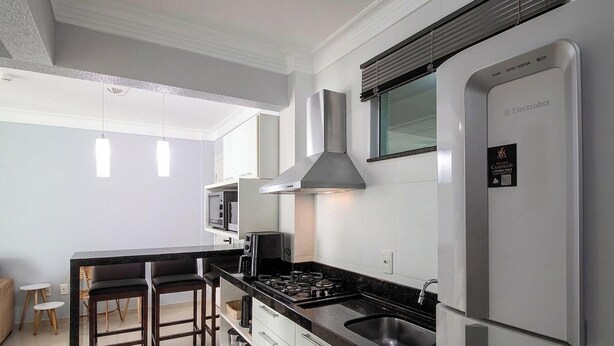 Cozinha moderna com fogão a gás, micro-ondas e exaustor de aço inoxidável. Há banquetas e uma janela com persianas.