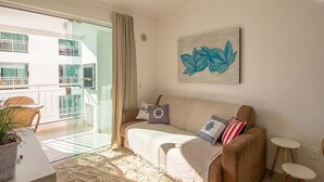 Uma sala de estar com sofá, uma mesinha, uma planta em vaso e uma varanda com vista para edifícios.